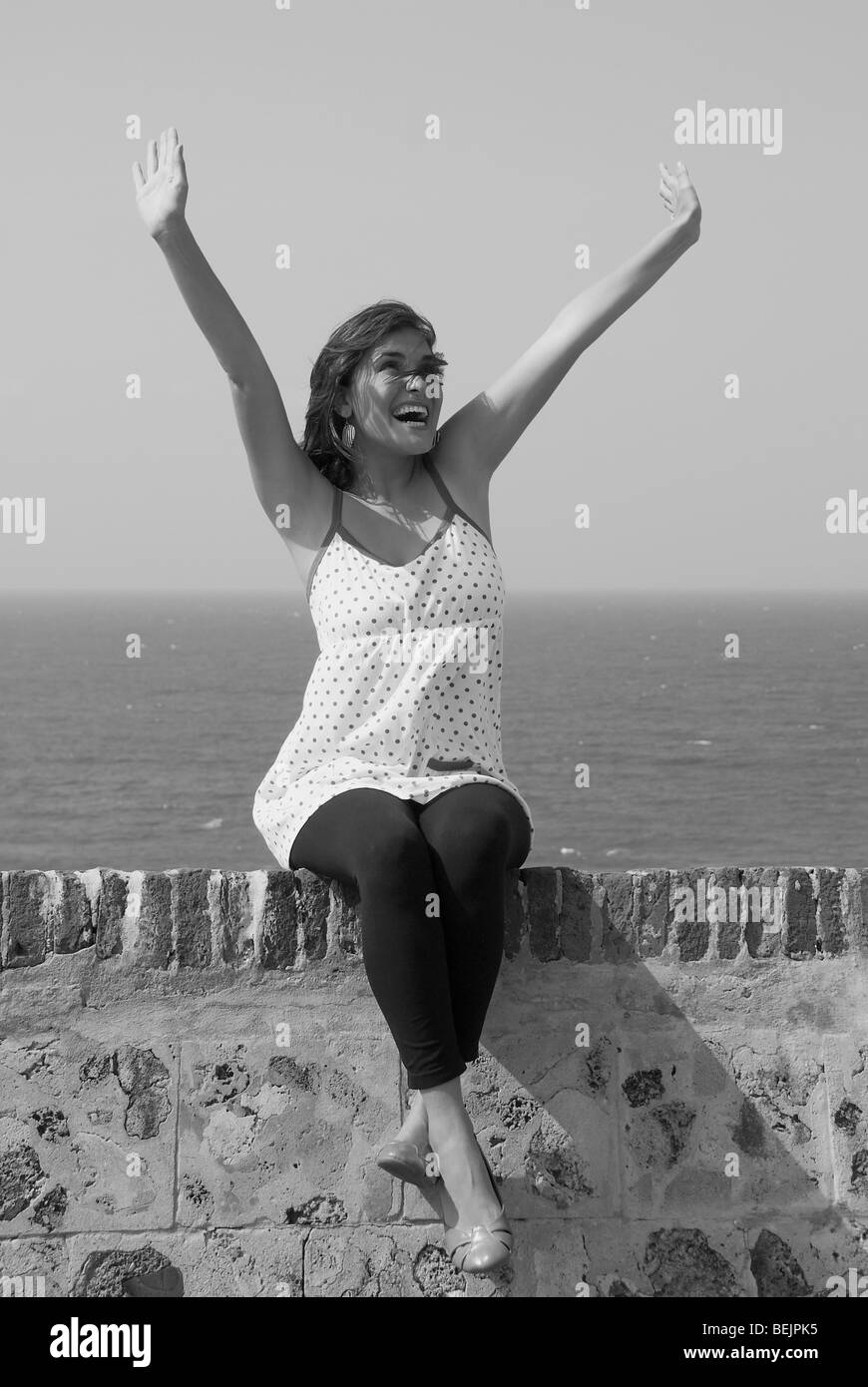 Young woman sitting on the ledge with her arms raised and laughing ...