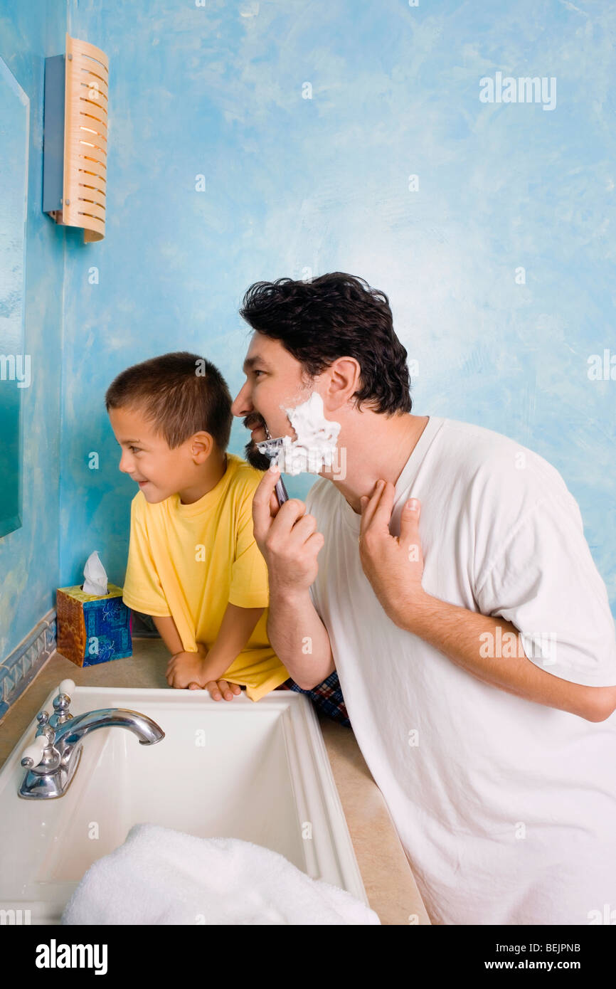 Side profile of a mid adult man shaving with his son sitting beside him ...