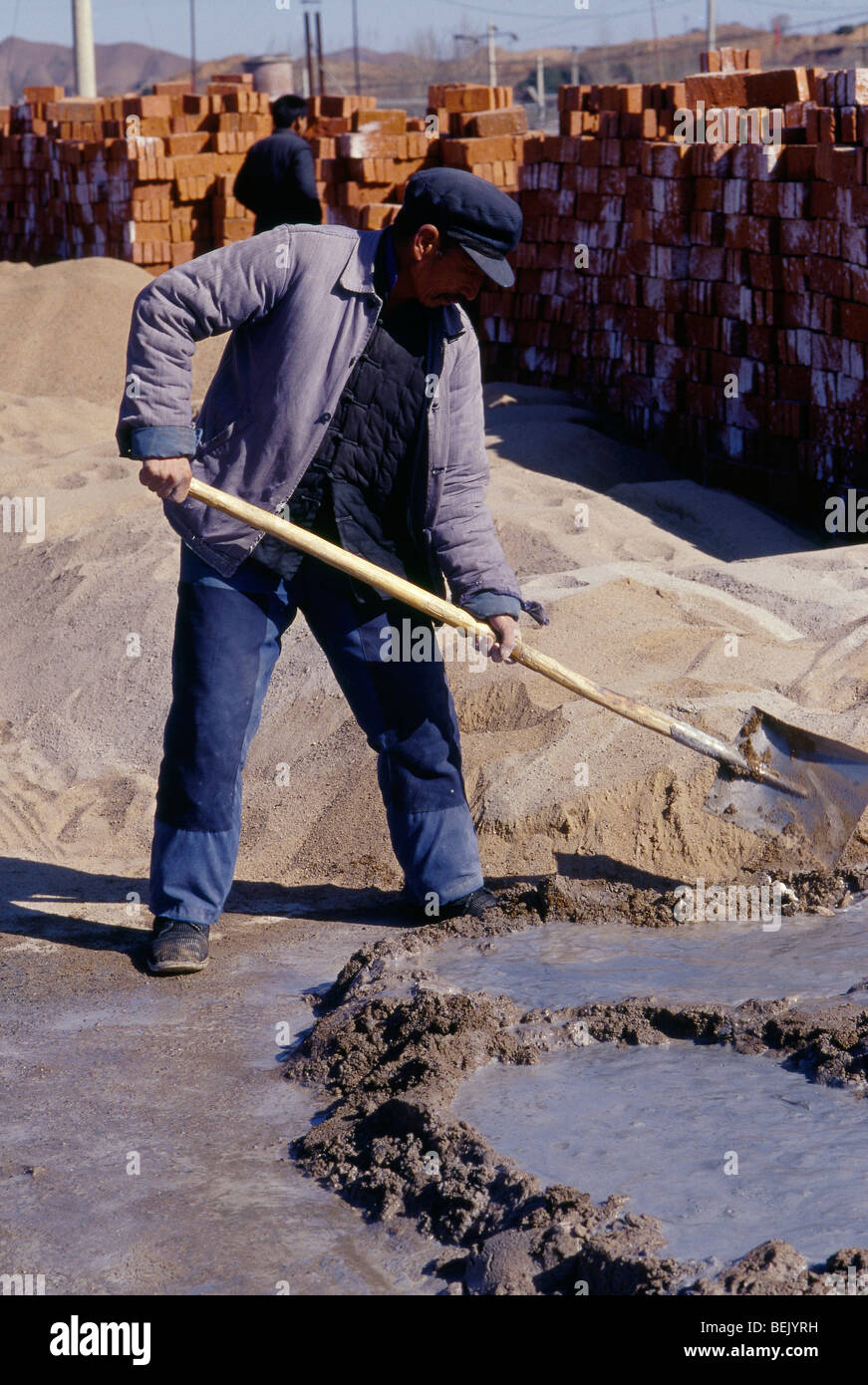 Male labourer at building site shovelling concrete and sand. Blue hat ...