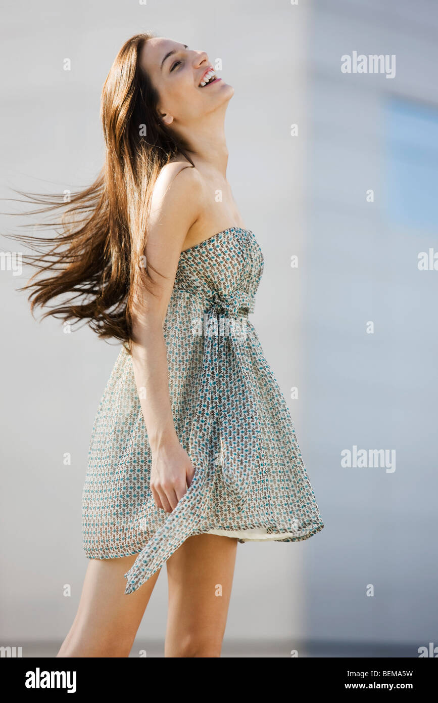 Young woman in sundress standing outdoors, laughing, hair tousled by ...