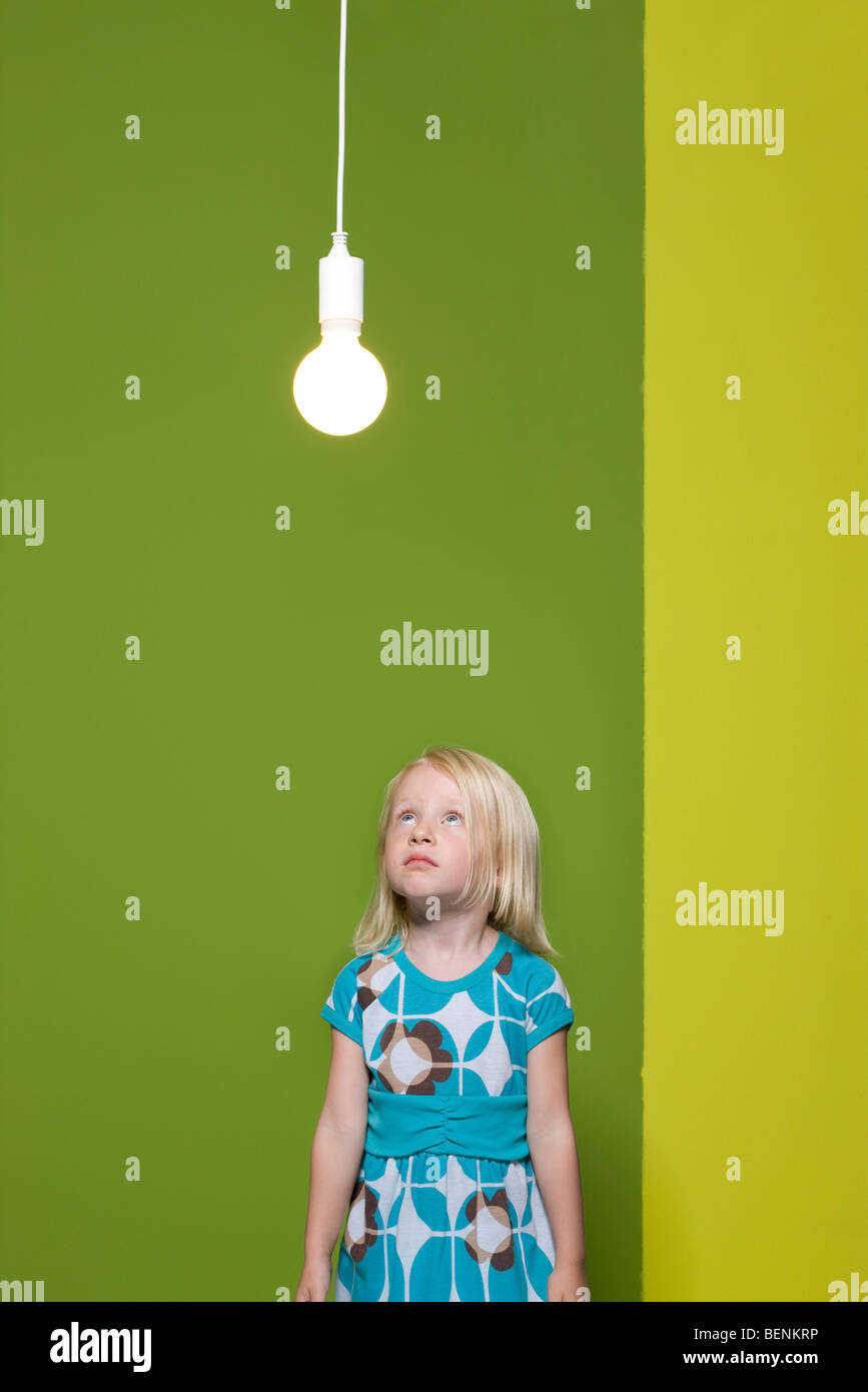 Little girl staring up at illuminated light bulb suspended overhead ...