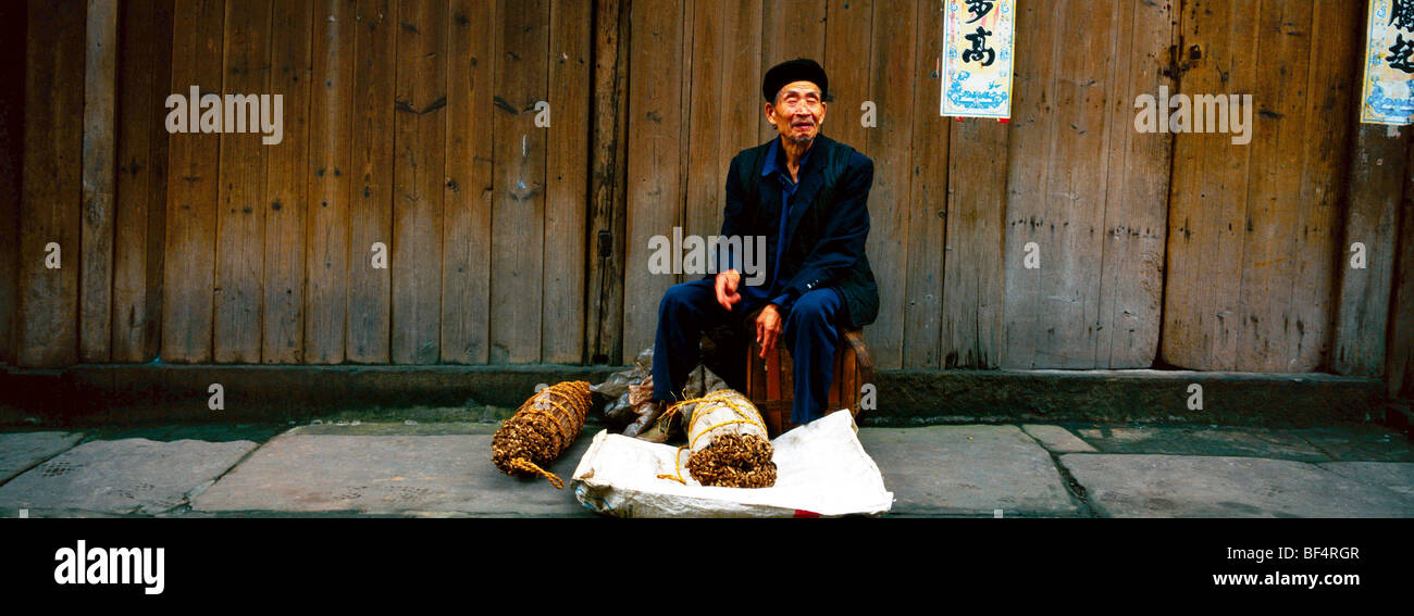 Elderly Chinese man selling tobacco leaves on the side of a stone paved ...