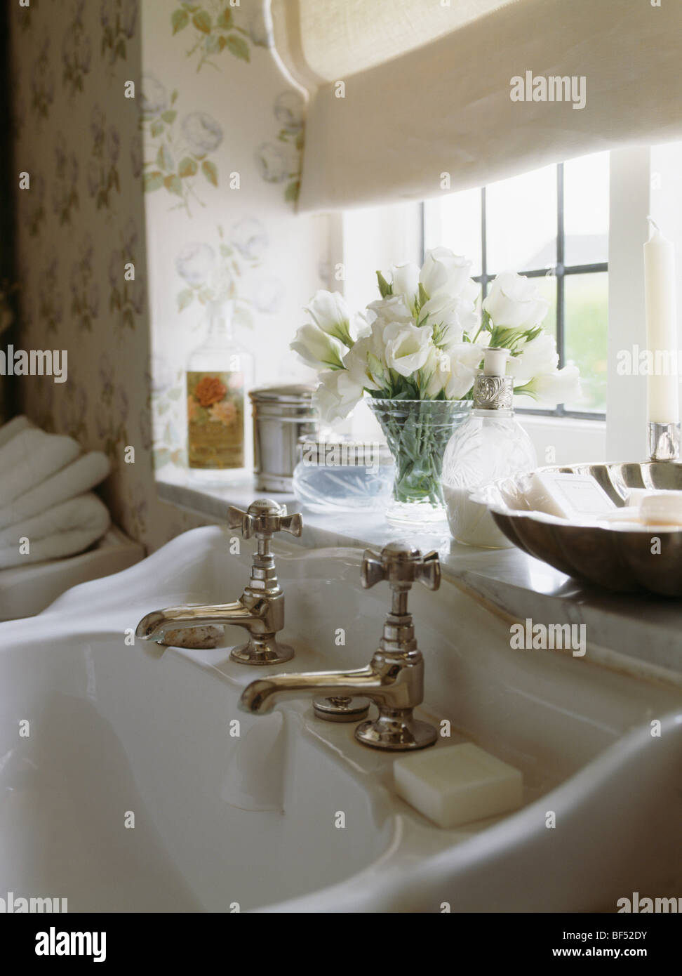 Close-up of brass taps on porcelain basin below window with vase of ...