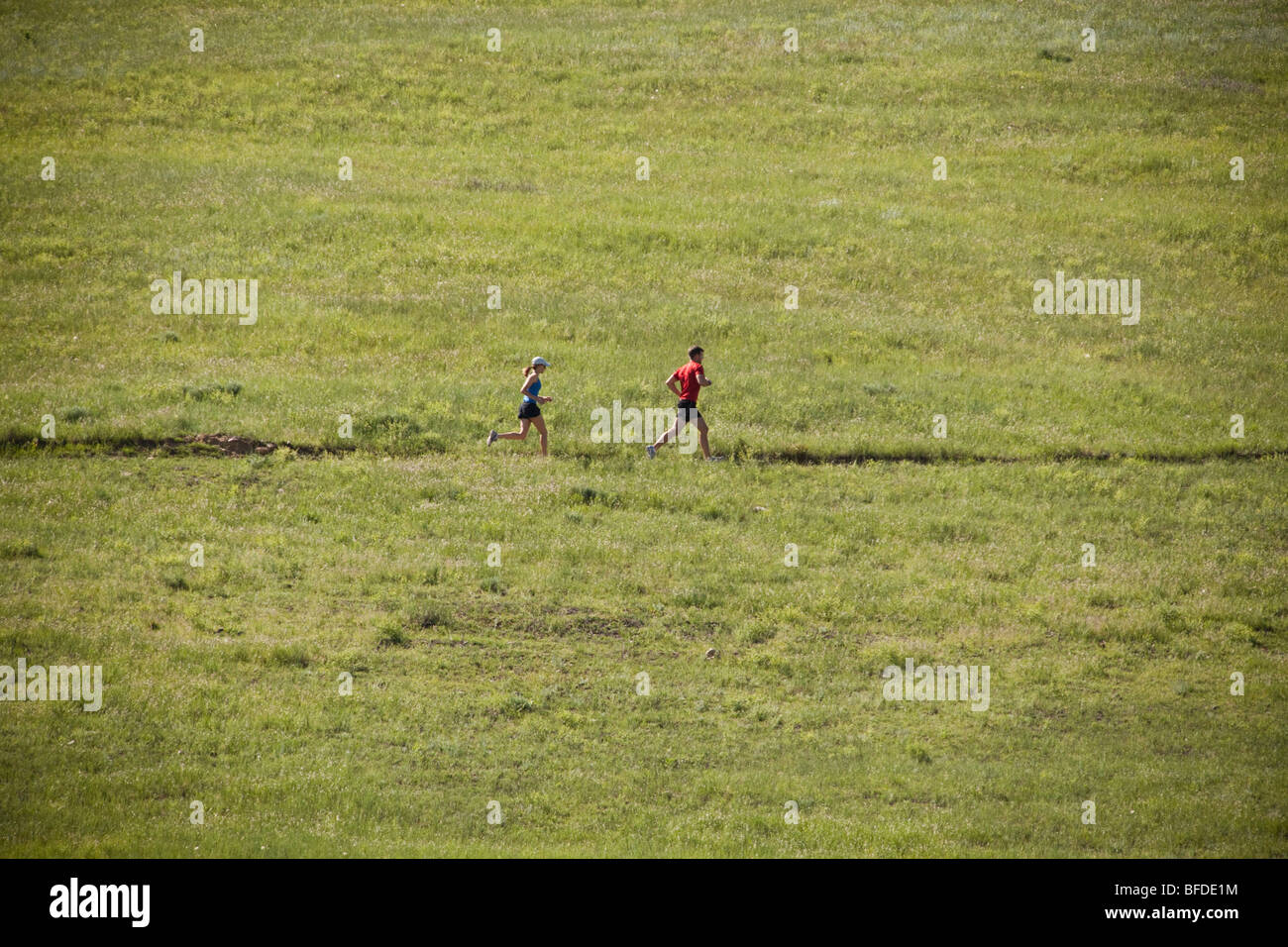 A man and woman run on a cut trail through the grassy foothills of the ...