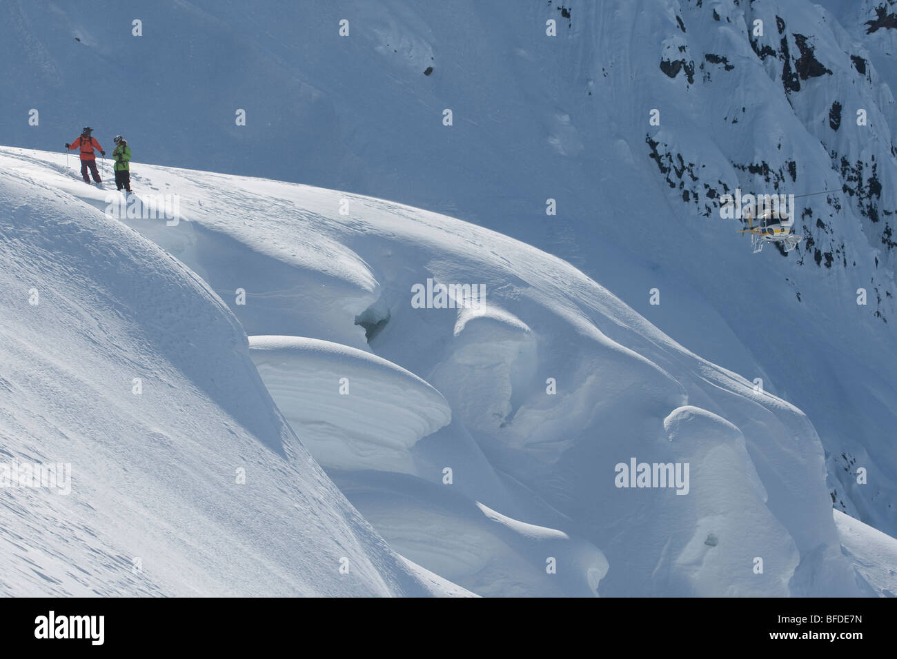 Two skiers waiting to ski with a helicopter in the background in Alaska ...