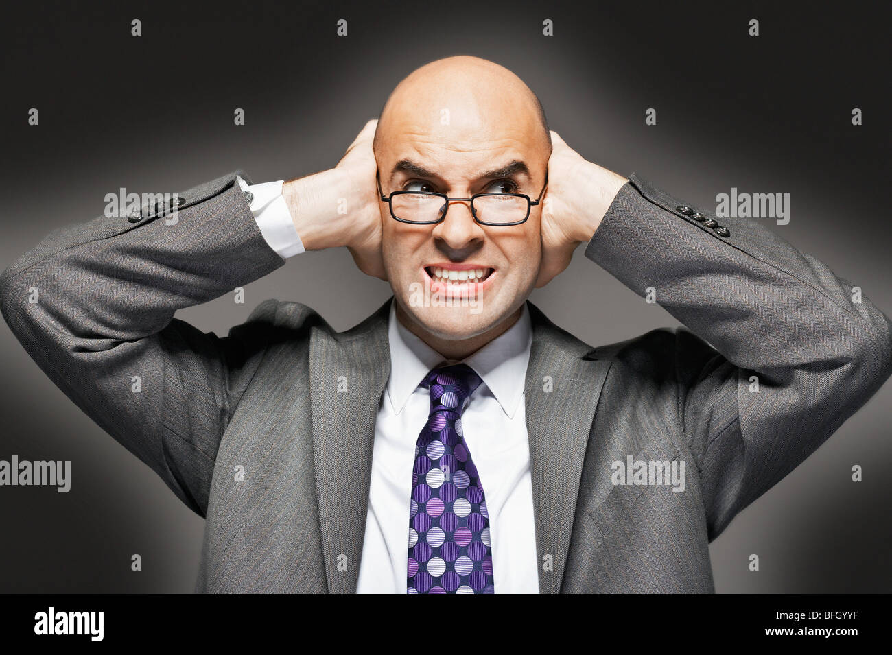 Bald businessman with hands over ears looking sideways against gray ...