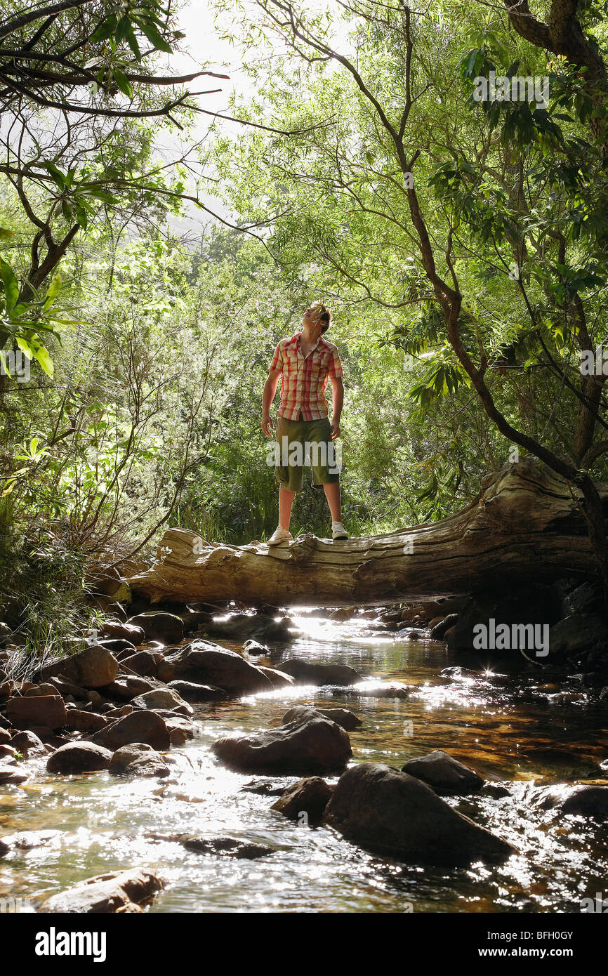 Teenage boy (16-17 years) standing on tree trunk above stream, looking ...