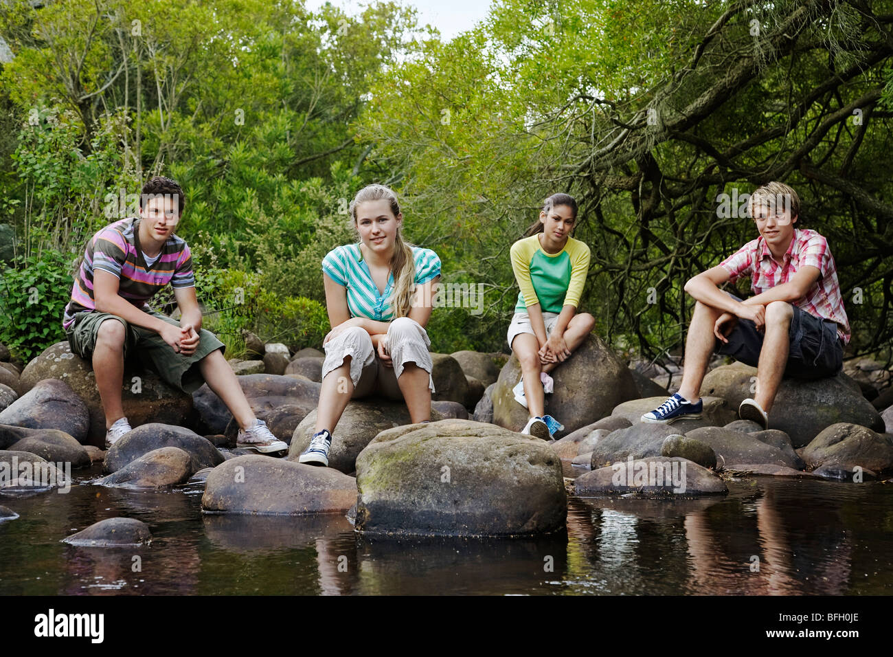 Portrait of four teenagers (16-17 years) sitting on stones by river ...