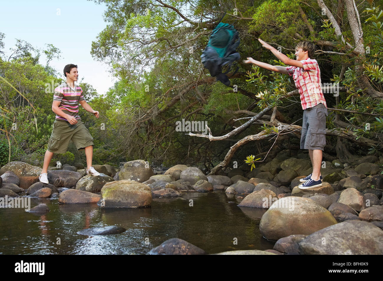 Two teenage boys (16-17 years) standing on stones by river, throwing ...