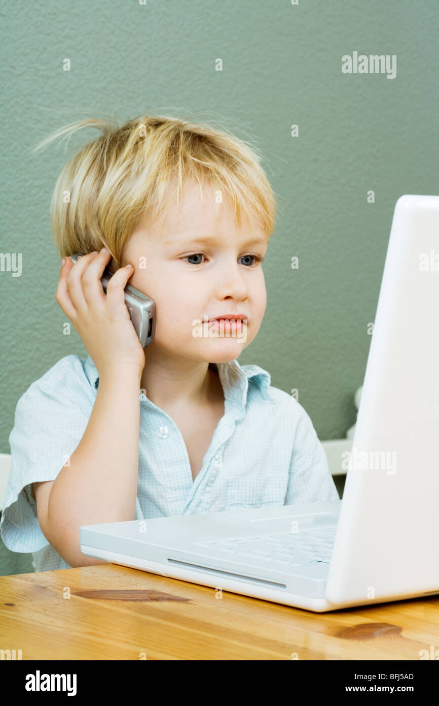 Scandinavian little boy using a mobile phone in front of a laptop ...
