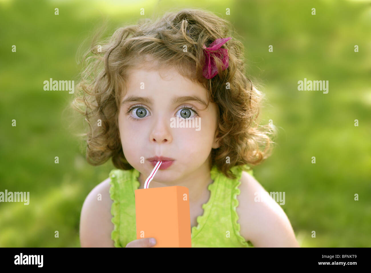 Little girl drinking juice from an orange brik on the park green grass ...