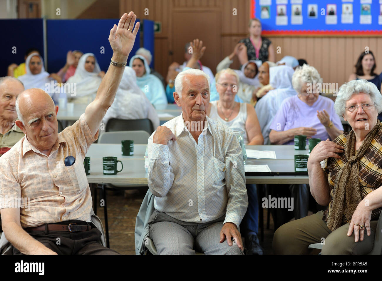 A mixed multi-cultural group of elderly people exercise in their chairs ...