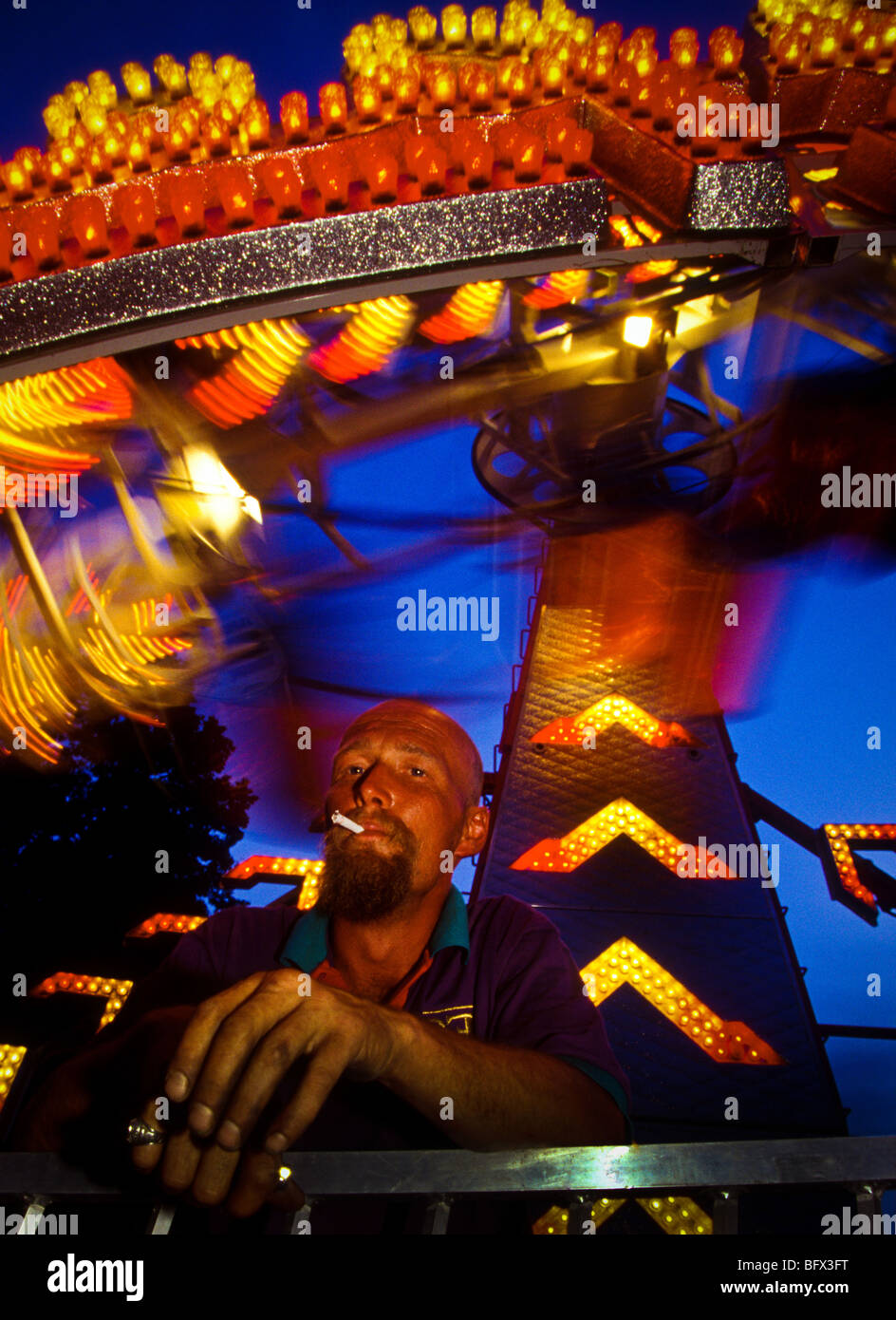 A carnival worker smokes as an amusement ride swings behind him Stock ...