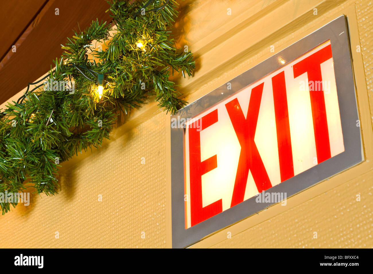 A closeup of an exit sign, lit, with green garland and white Christmas ...