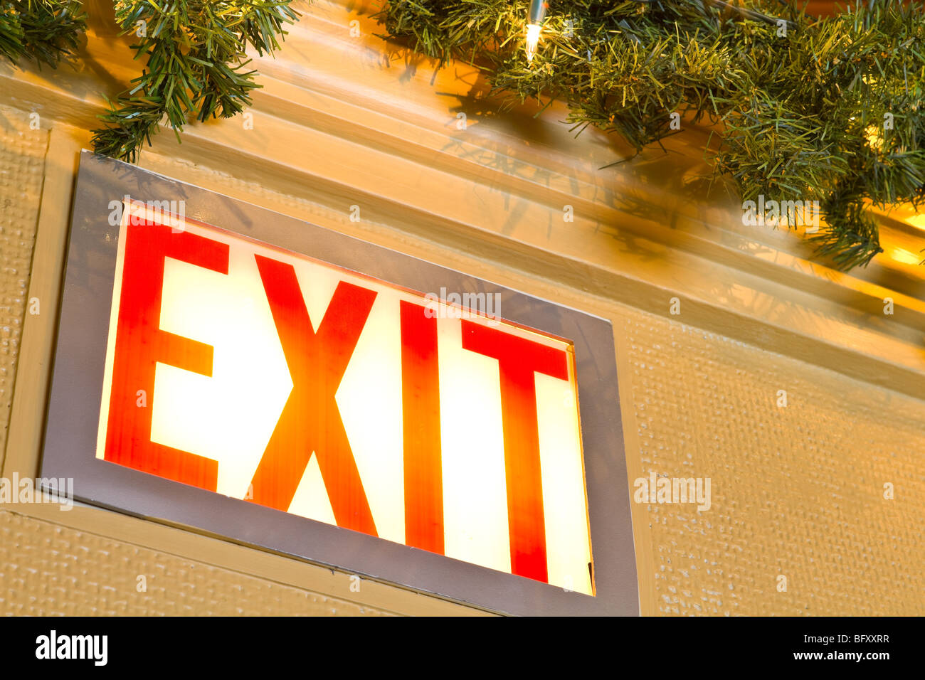 A closeup of an exit sign, lit, with green garland and white Christmas ...