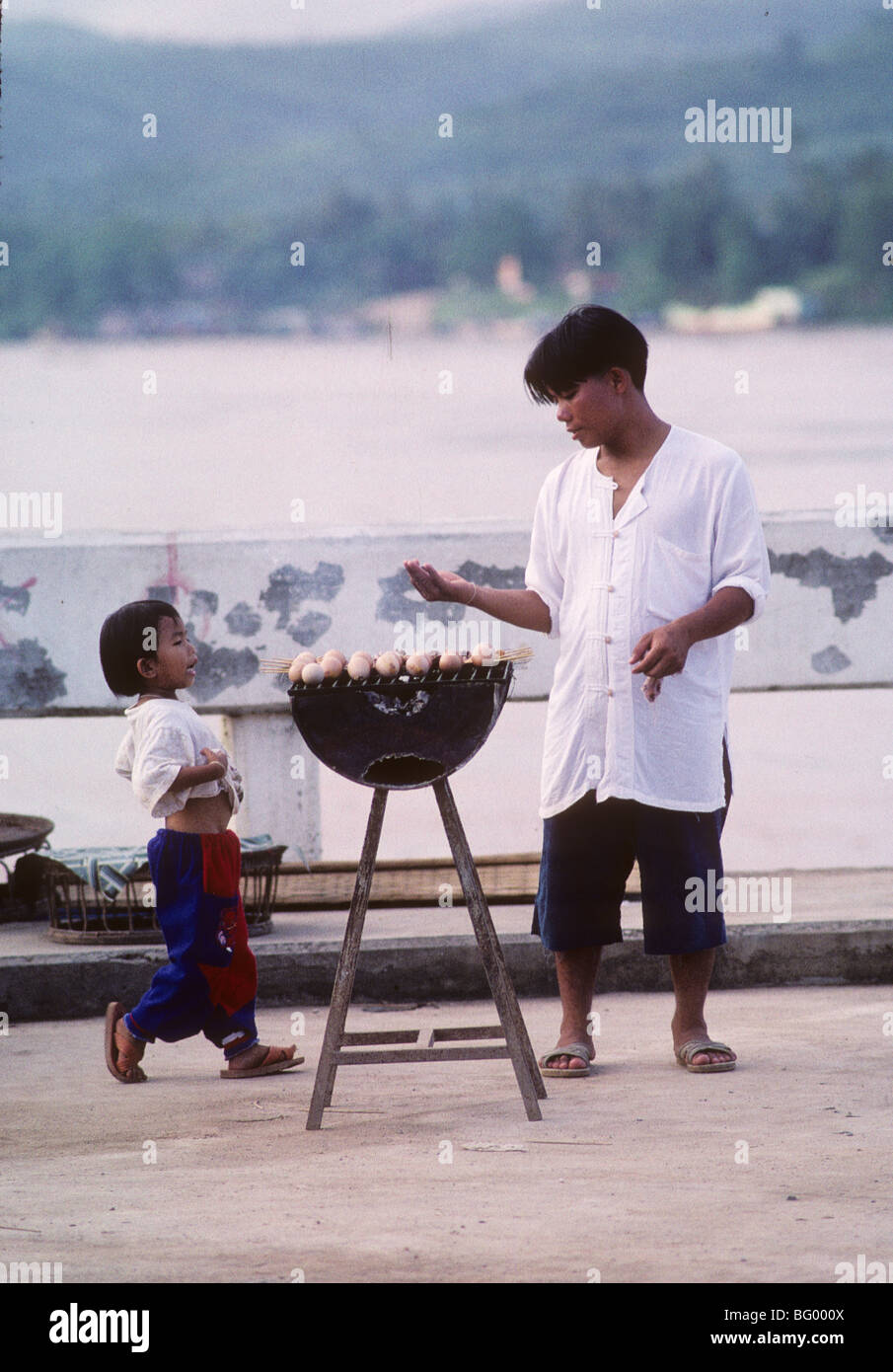 A young boy watches his father barbecue eggs at the Mekong River in ...