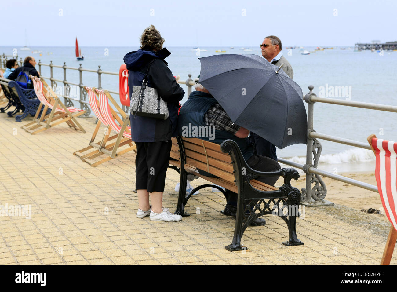 Two people sitting on seats at the seaside using an umbrella to shelter ...