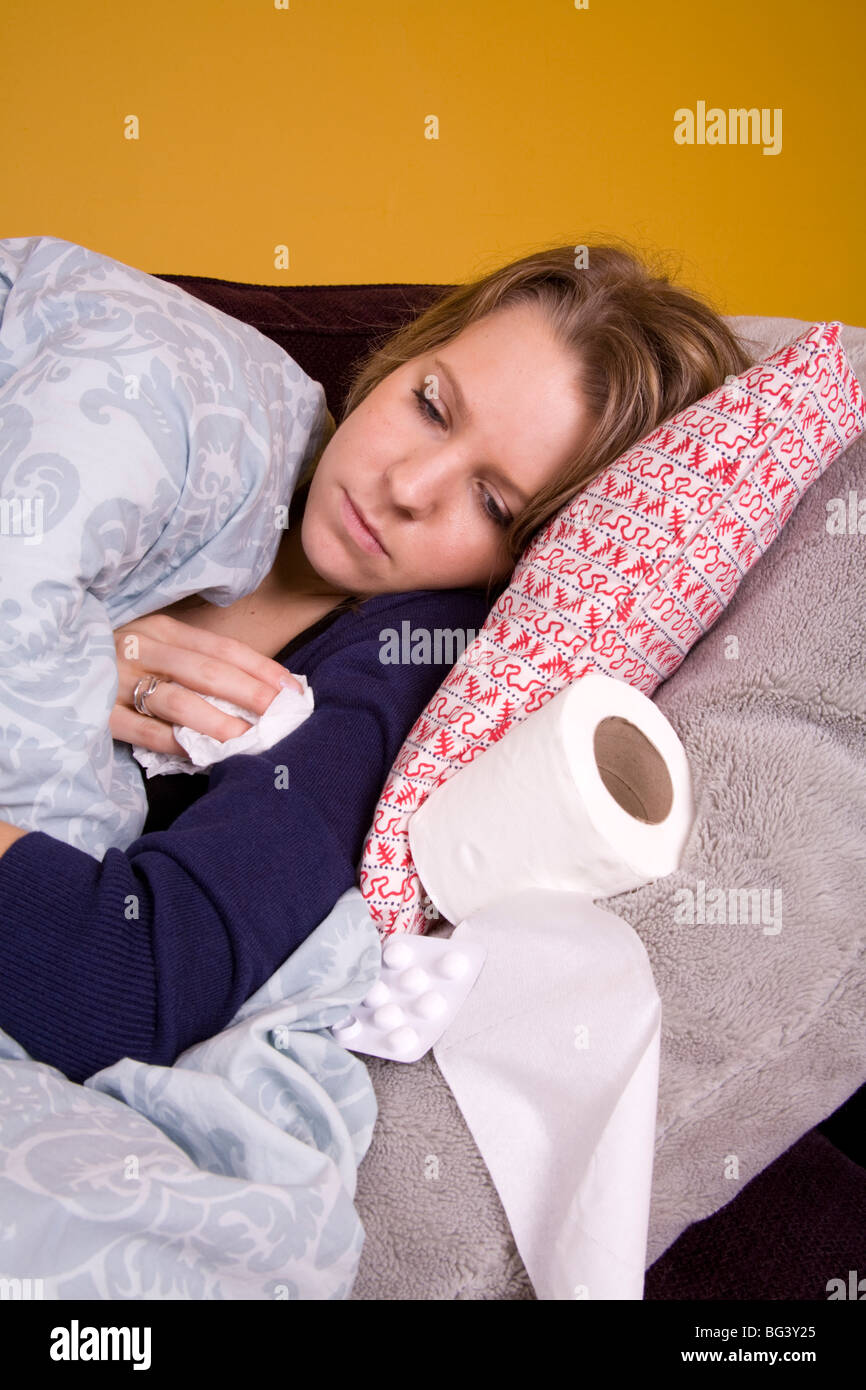 Young woman suffering from a cold or flu, curled up under a duvet, with ...