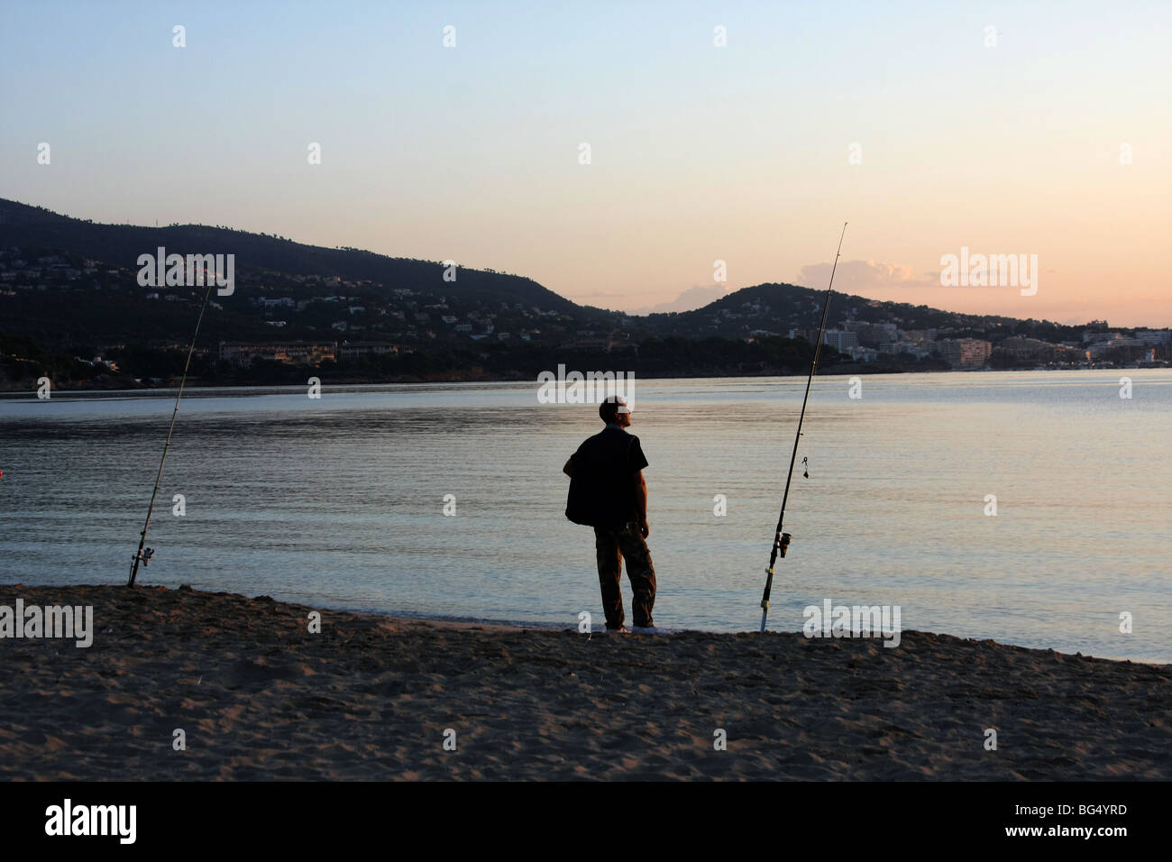 Beach angler in silhouette against calm sea in light just before ...