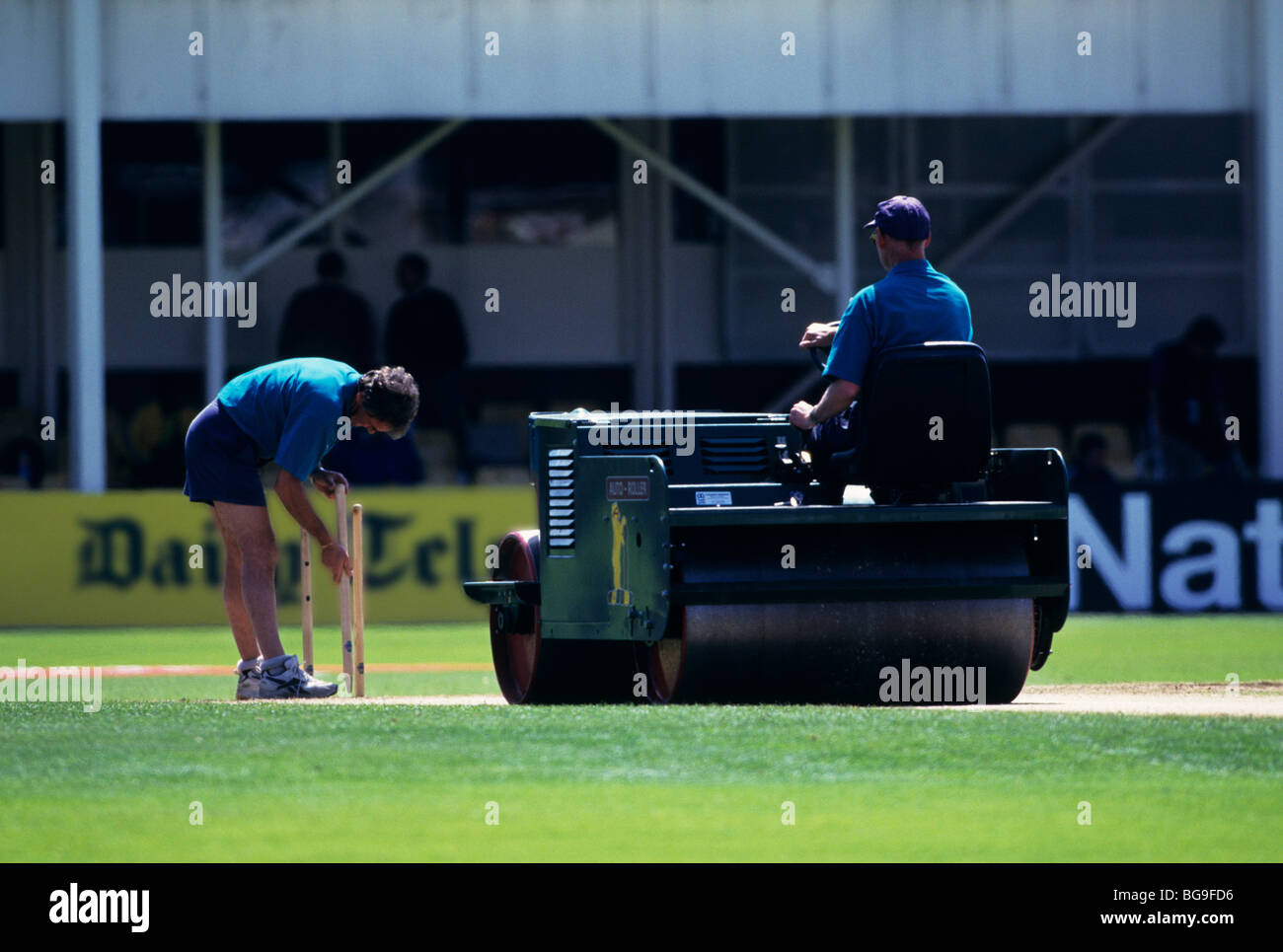 Man driving a bowdry over a cricket pitch whilst groundsman places ...
