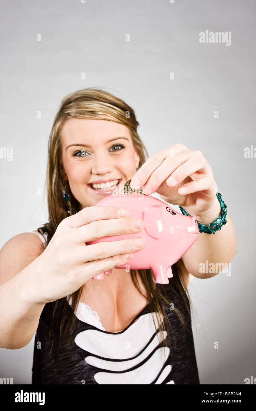Pretty young woman smiling and putting coin into a pink piggy bank ...