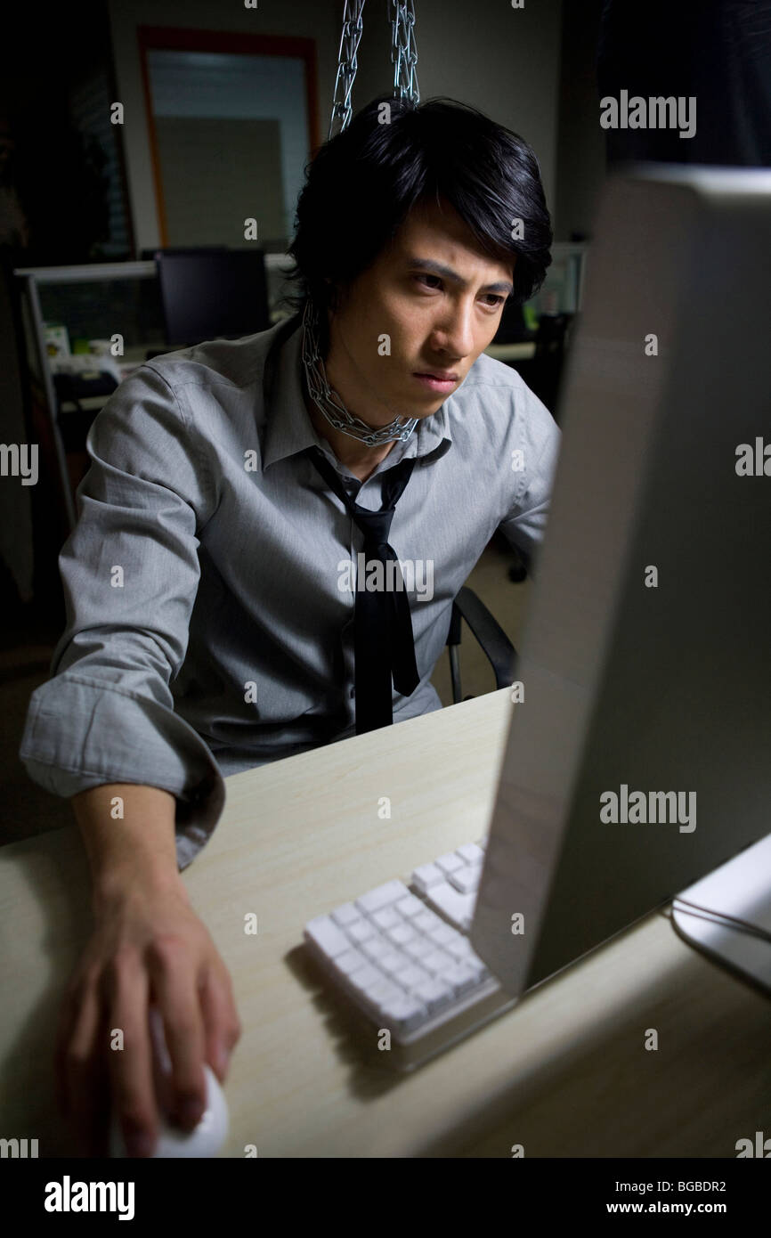 Young man in shirt and tie is hung from a chain at his cubicle while he ...