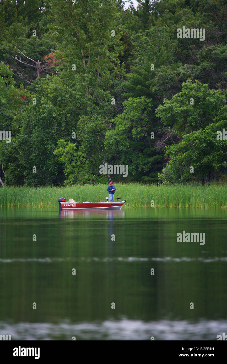 Fisherman in a red Lund fishing boat fishing along grassy shoreline ...