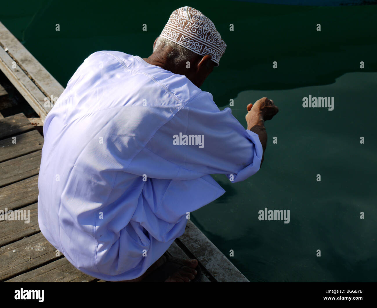 Omani man in traditional dress fishing from a pier near the Muscat fish ...
