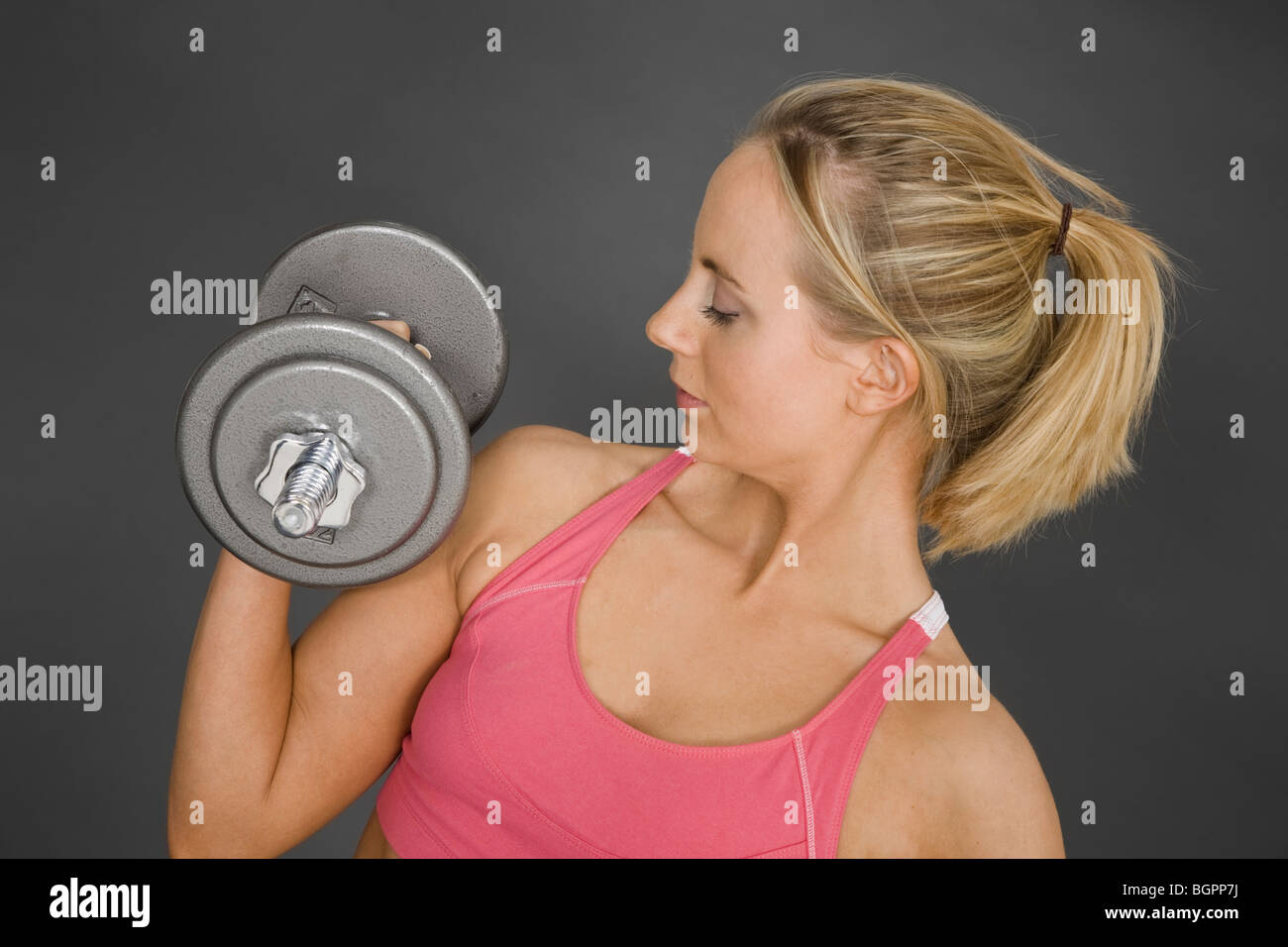 Caucasian woman in early 20s lifting weights using a dumbbell on a gray ...