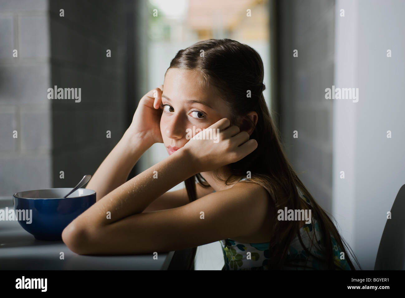 Girl sitting at table with head in hands, looking sulkily at camera ...