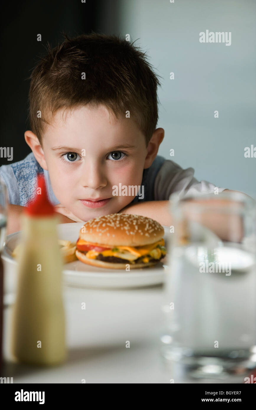 Little boy sitting at table with cheeseburger on plate, resting head on ...