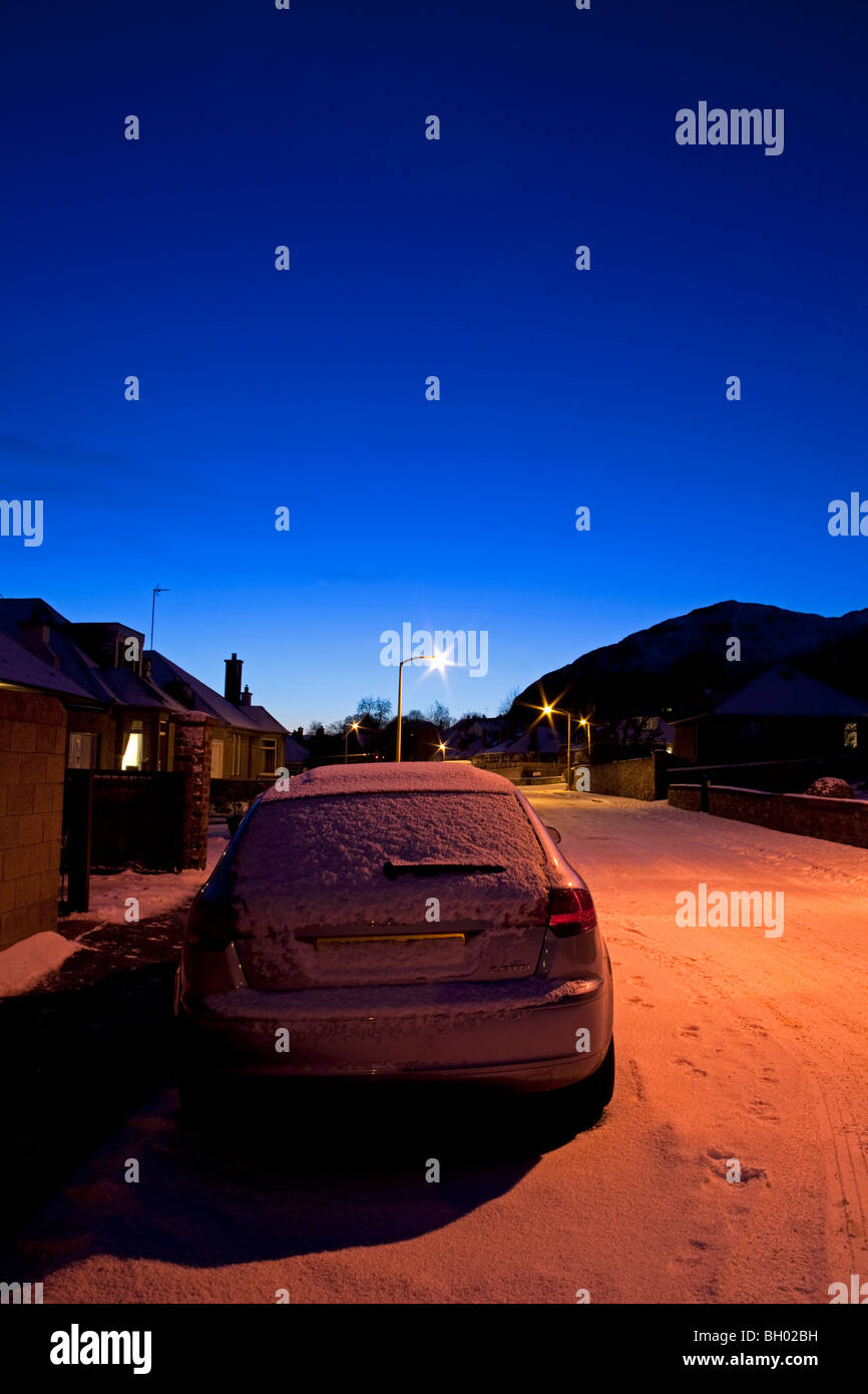 Parked car covered in winter snow under sodium street lighting at dusk ...