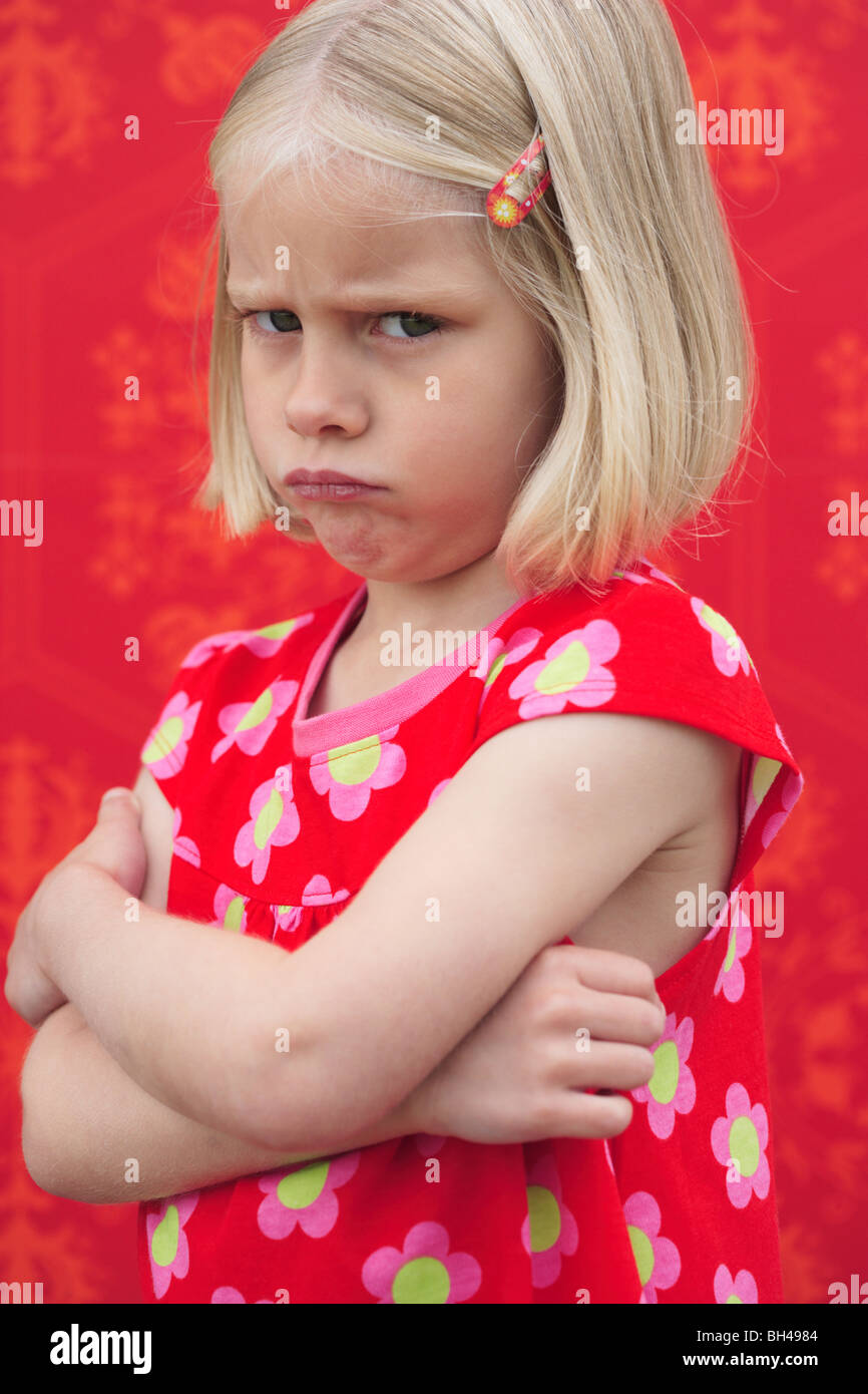 Young girl wearing a red dress standing with her arms folded frowning ...
