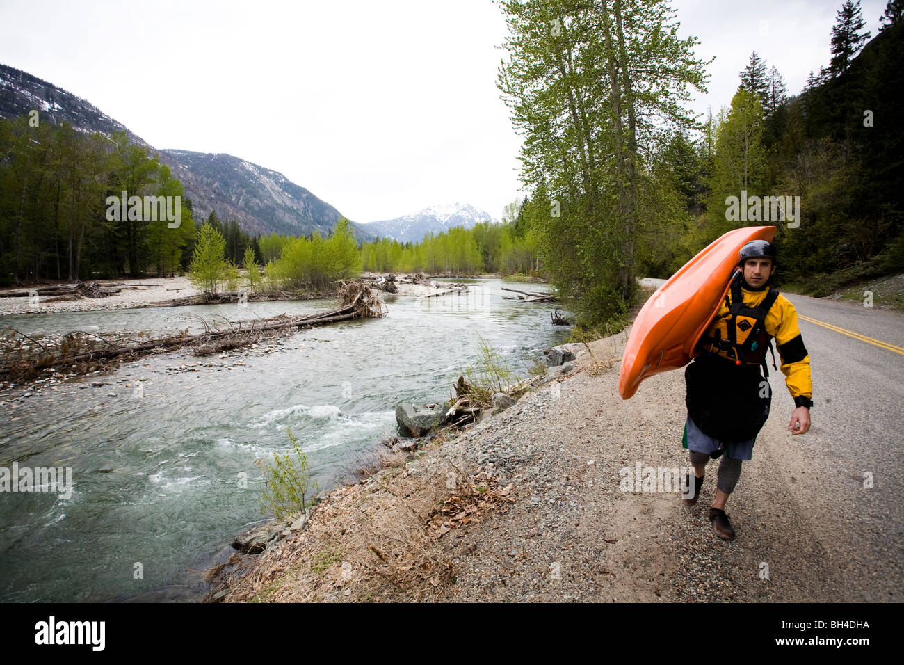 A young man carries a kayak down a road with a river alongside him ...