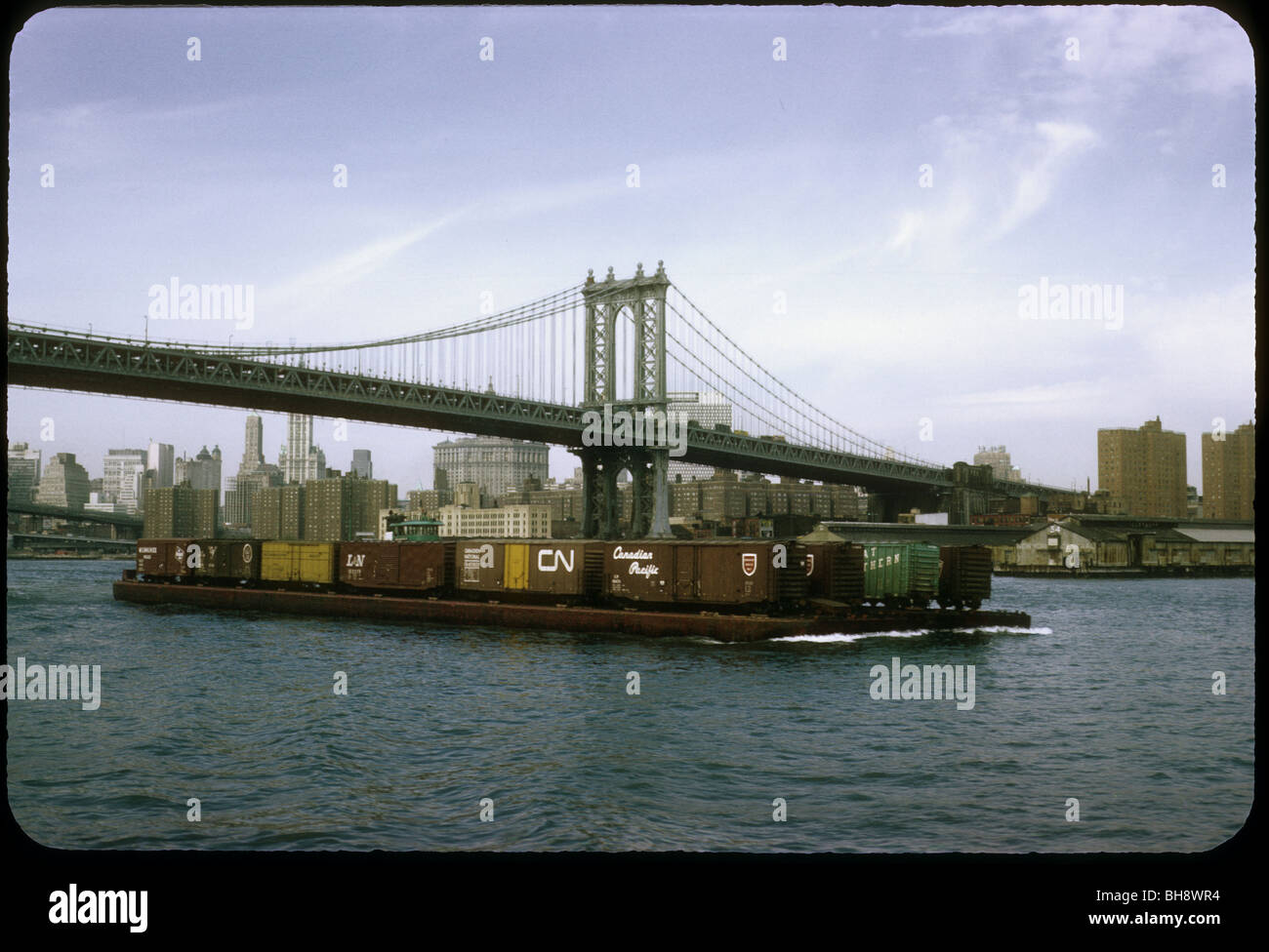 A barge takes boxcars. 1968. Manhattan Bridge crosses the East River ...