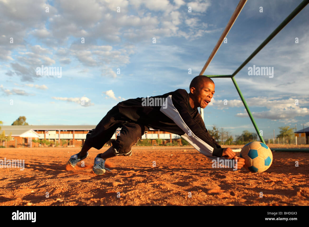 A soccer goalkeeper dives to save the shot at goal on a sandy pitch in ...