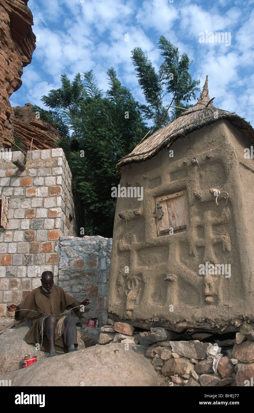 A Dogon man making rope beside a decorated mud hut, below the ...