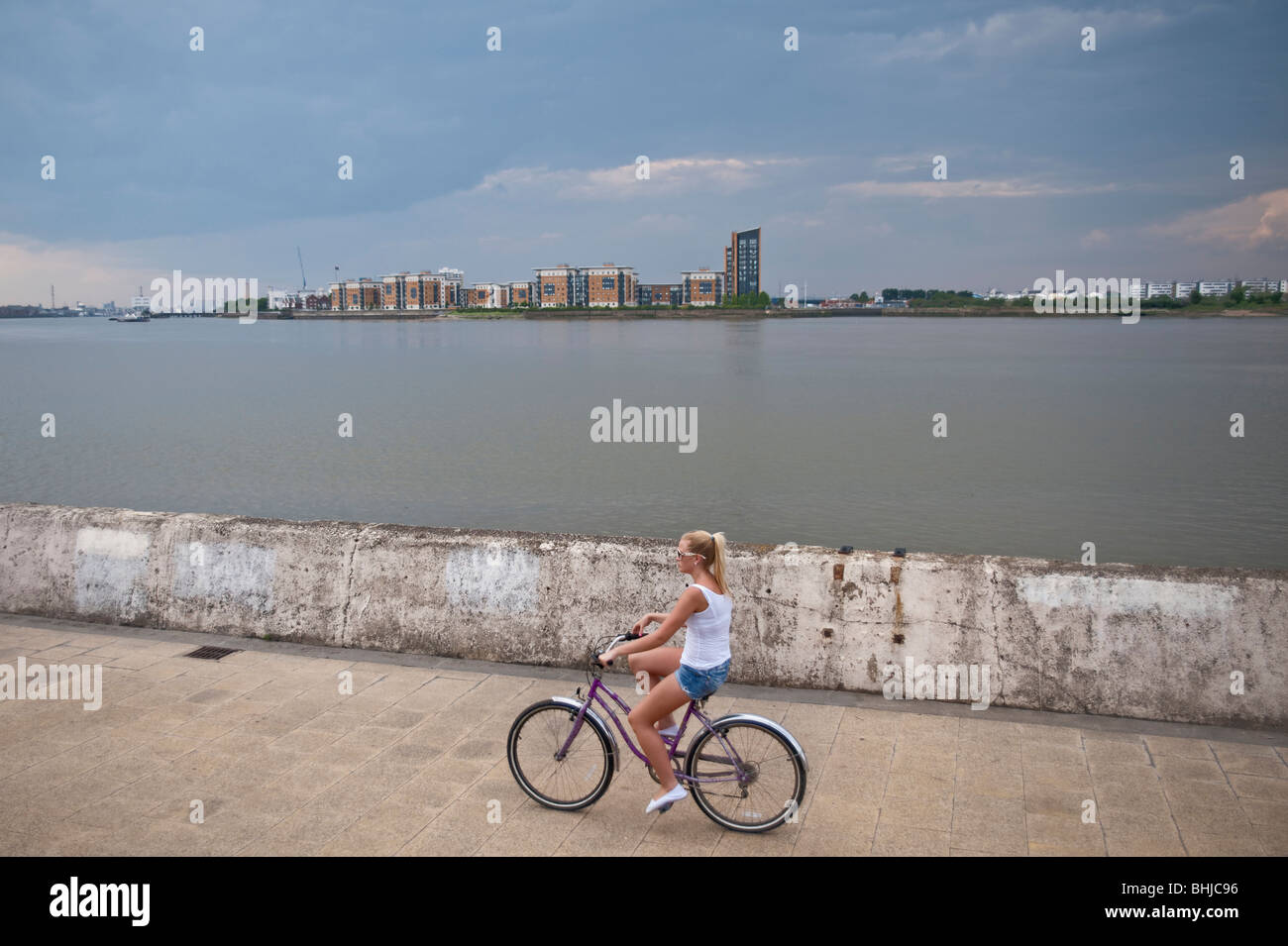 Woman rides bicycle along Thames path at Thamesmead with view across ...