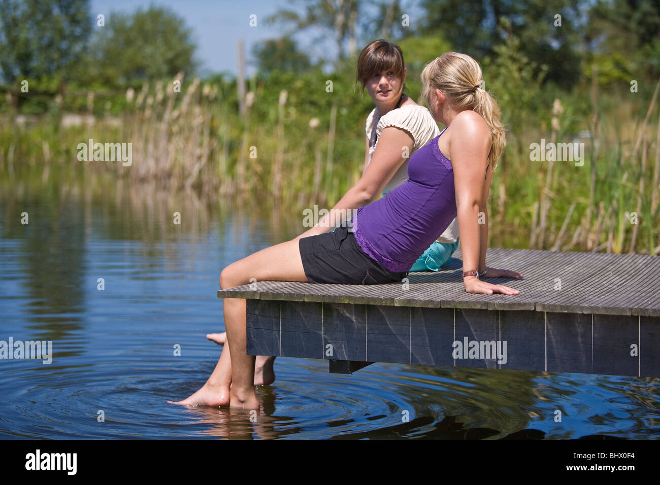 Two young women sitting at a lake talking Stock Photo - Alamy