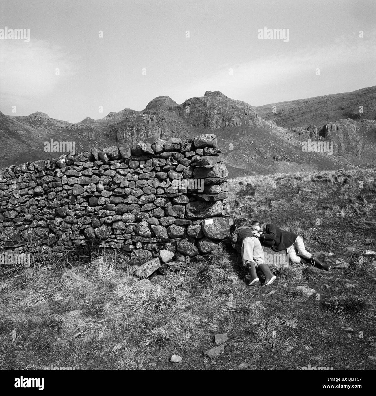 Two children lay down on the ground at the foot of a solid dry stone ...