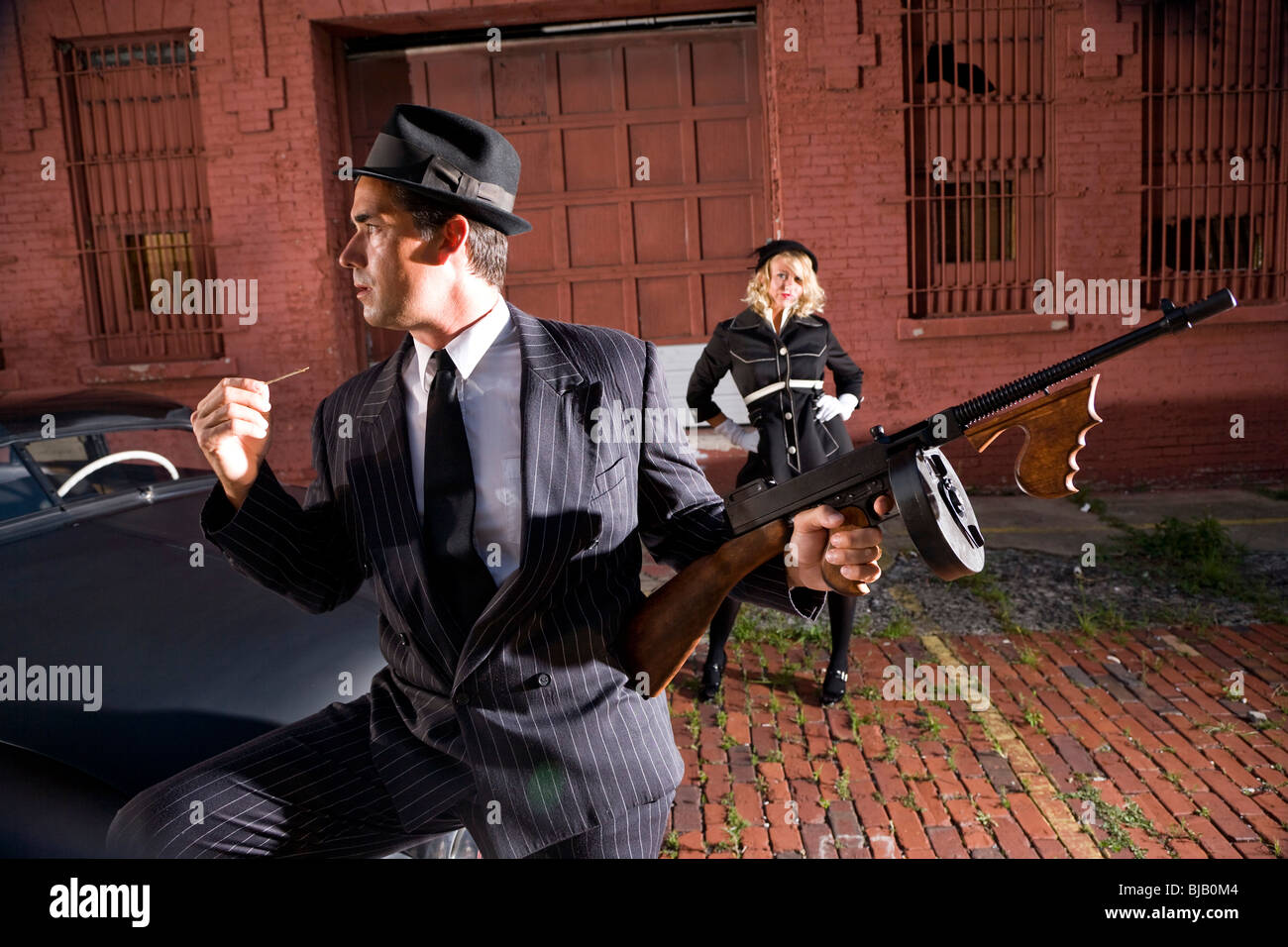 Gangster holding Tommy gun leaning on vintage car, girl in background ...