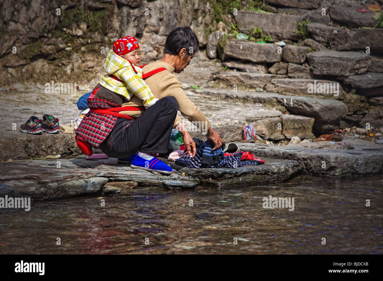 A village woman washing clothings at a stream, with her baby on her ...