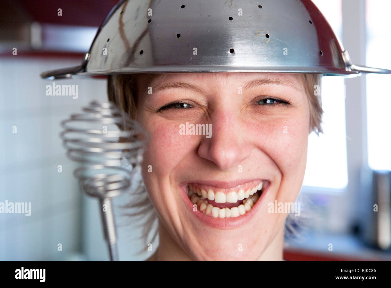 Smiling woman in a kitchen with a kitchen sieve on her and a whisk in ...