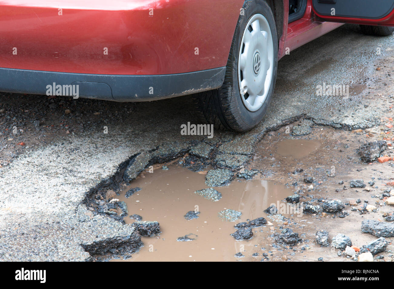 car and wheel beside a pothole left in road by harsh winter weather ...