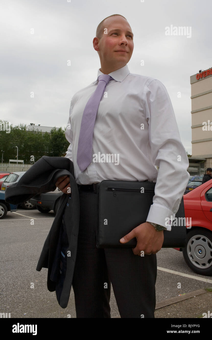 A salesman in his 20's stands in a car park holding his suit jacket ...