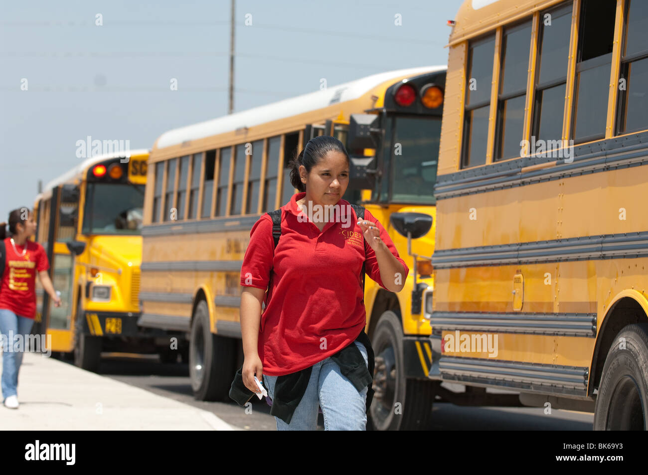 Female Hispanic high school student walks along row of yellow school ...