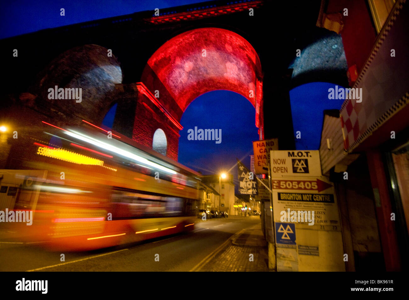 A bus speeds under an old railway viaduct arch illuminated at night by ...