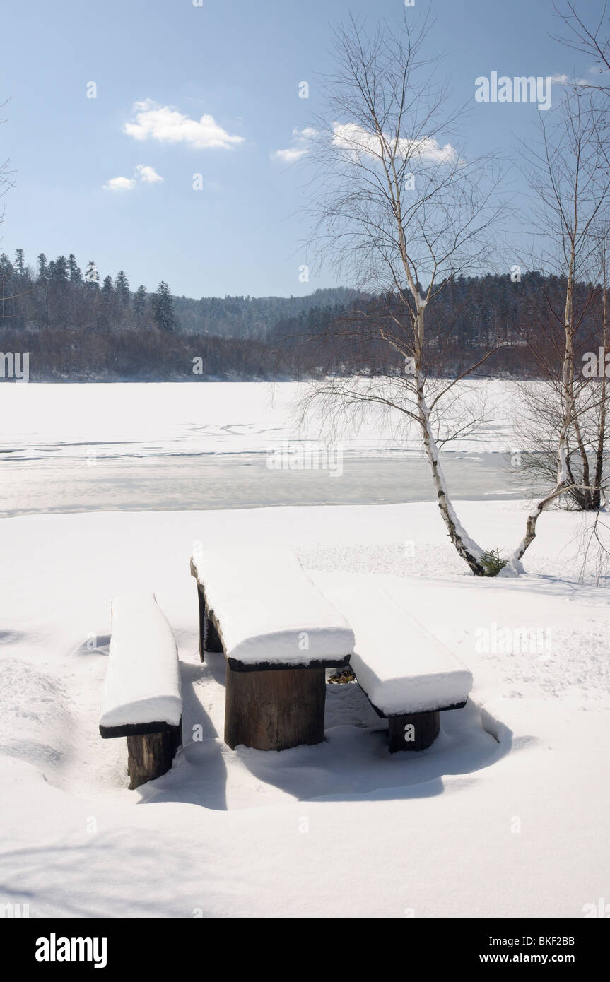 Wooden picnic table beside frozen lake, covered with snow during winter ...
