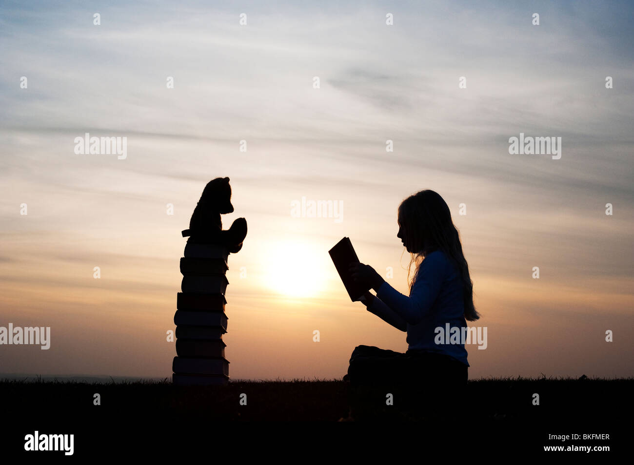 Young Girl sitting down reading a book opposite a teddy bear sitting on ...