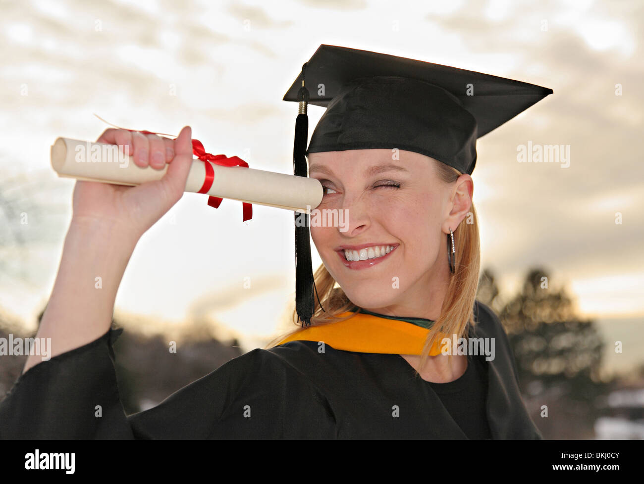 smiling female college graduate in cap and gown looking through rolled ...